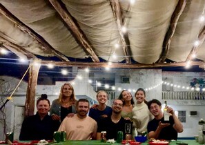Um grupo de pessoas sentadas à mesa, com comida e bebida, sob uma estrutura semelhante a uma tenda com luzes de fada.