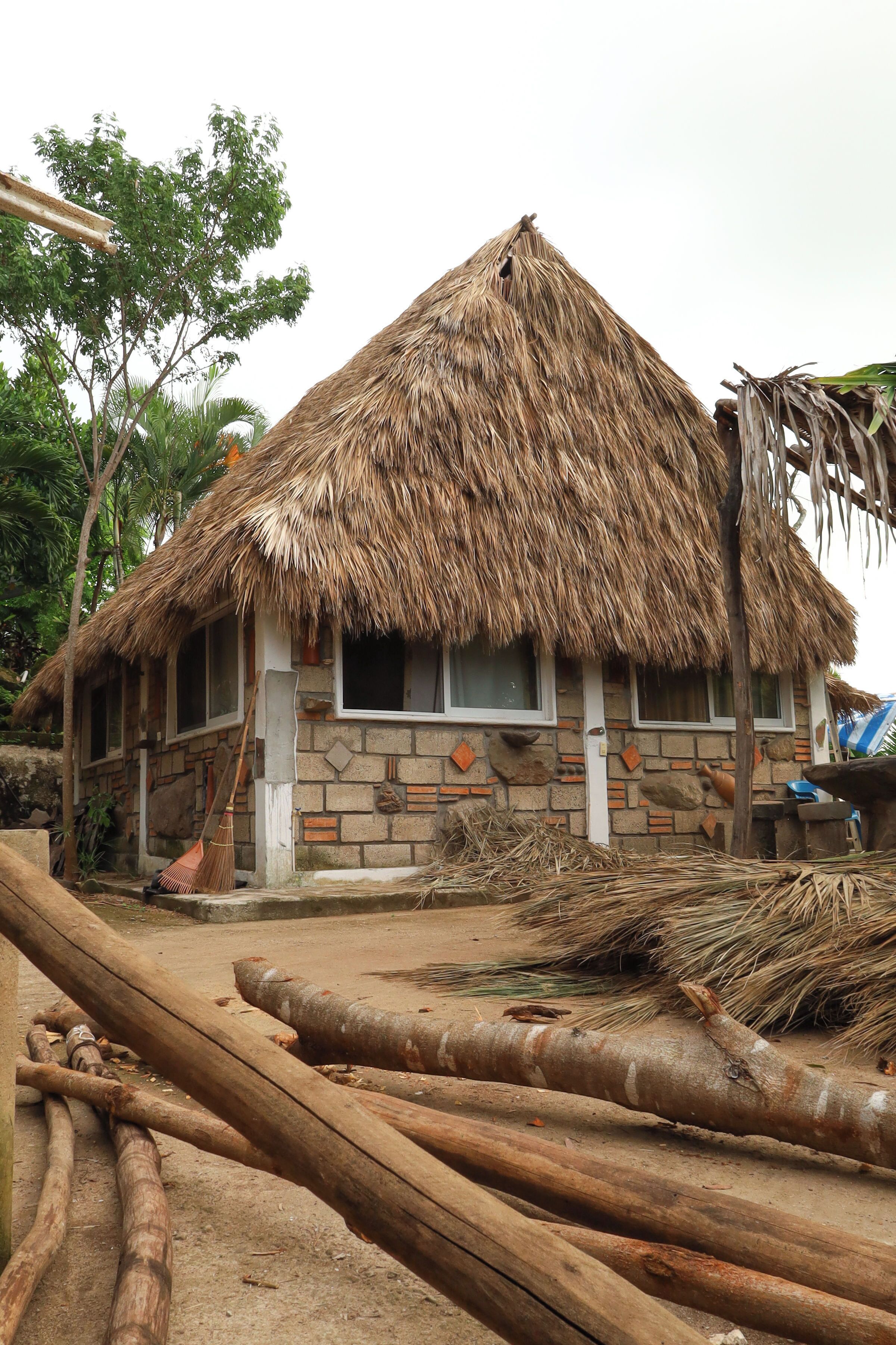 Uma casa rústica com telhado de palha e fachada de pedra, rodeada por vegetação natural e troncos de madeira.