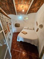 Um quarto pequeno e acolhedor, com teto de tijolos, uma cama com roupa de cama branca e uma janela pequena.