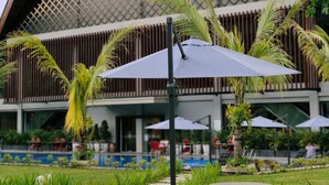 Edifício moderno com telhado inclinado e fachada em tela metálica. Na frente, área de estar com mesa e cadeiras, cercada por vegetação e piscina.
