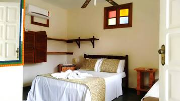 Quarto com cama, mesa de cabeceira, mesinha, prateleira na parede e janela com vitrais.