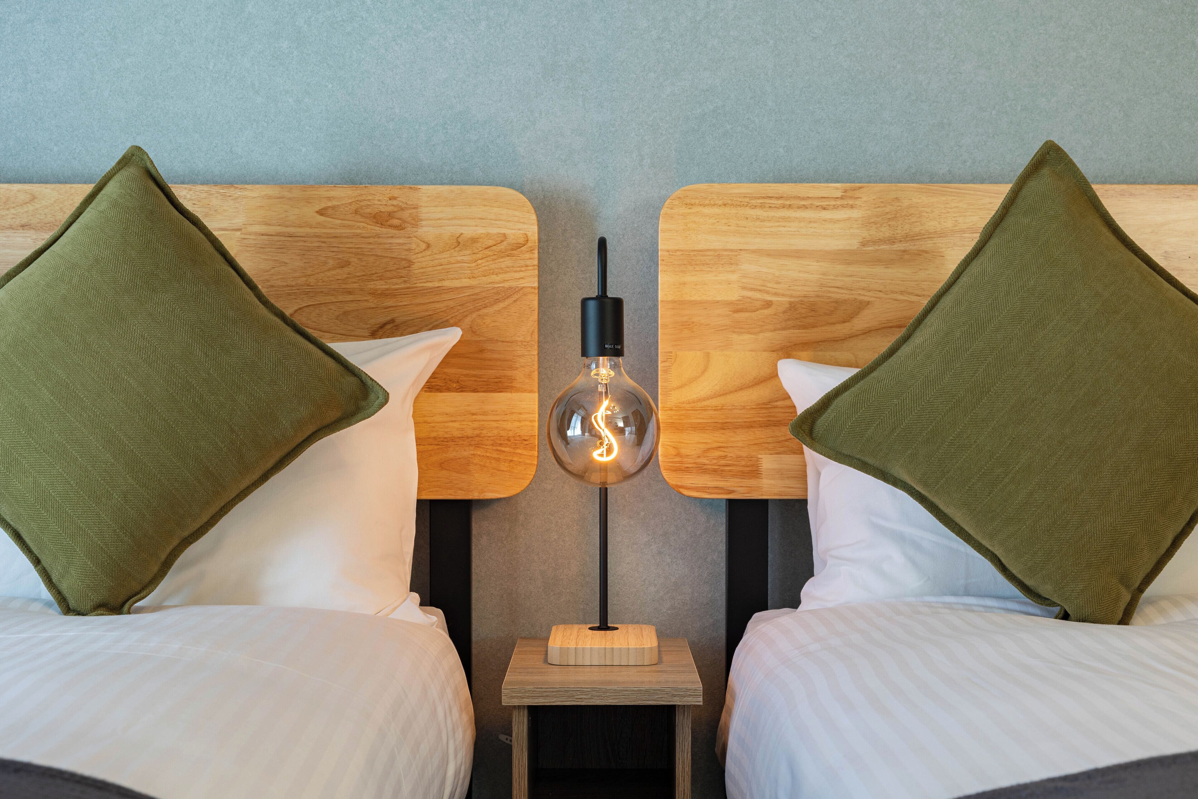 Quarto de hotel com duas camas, cada uma com cabeceira de madeira e um travesseiro verde. Uma pequena mesa de cabeceira de madeira com abajur fica entre as camas.