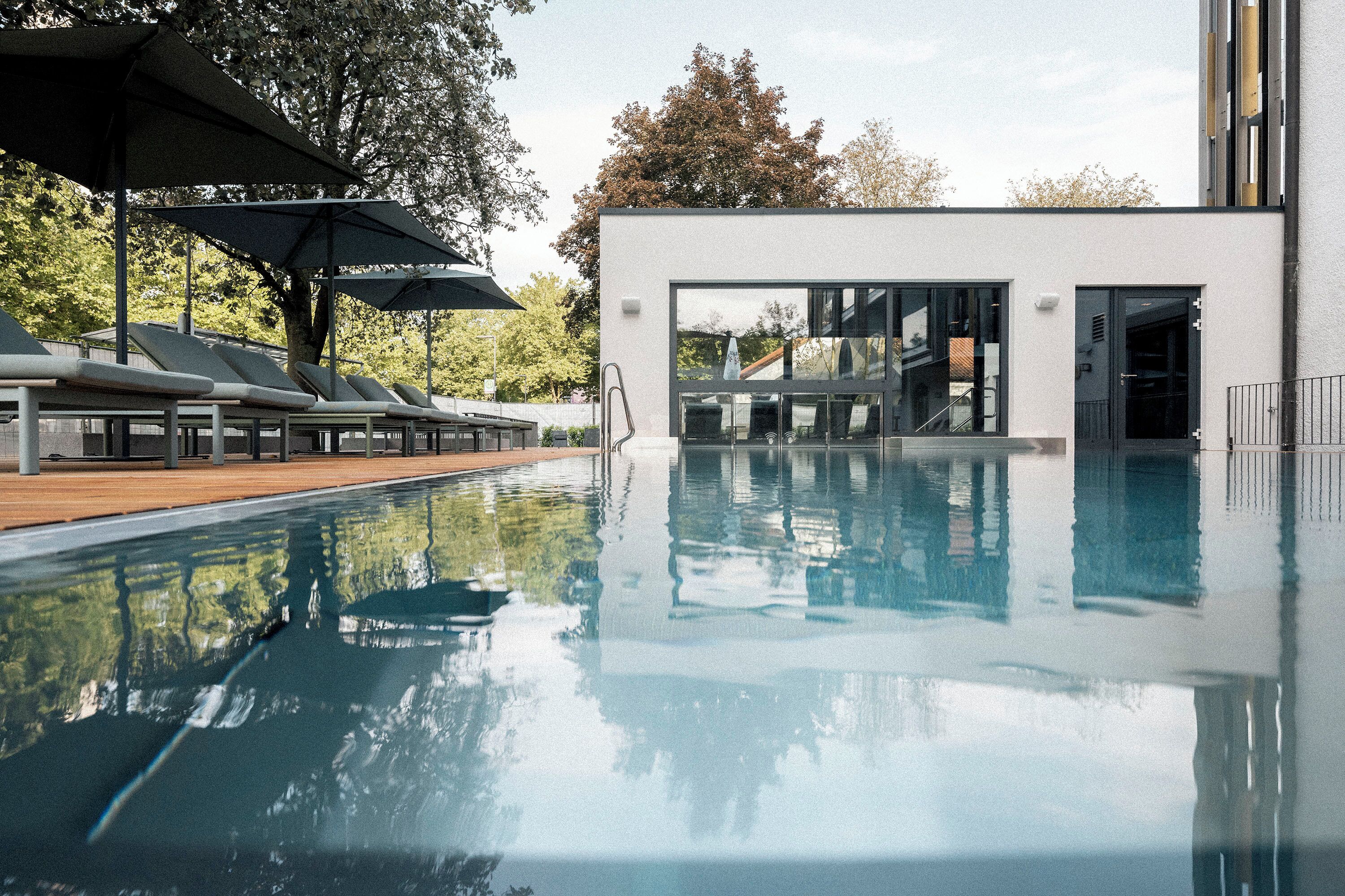 Uma área de piscina moderna com espreguiçadeiras e guarda-sóis, rodeada por árvores.