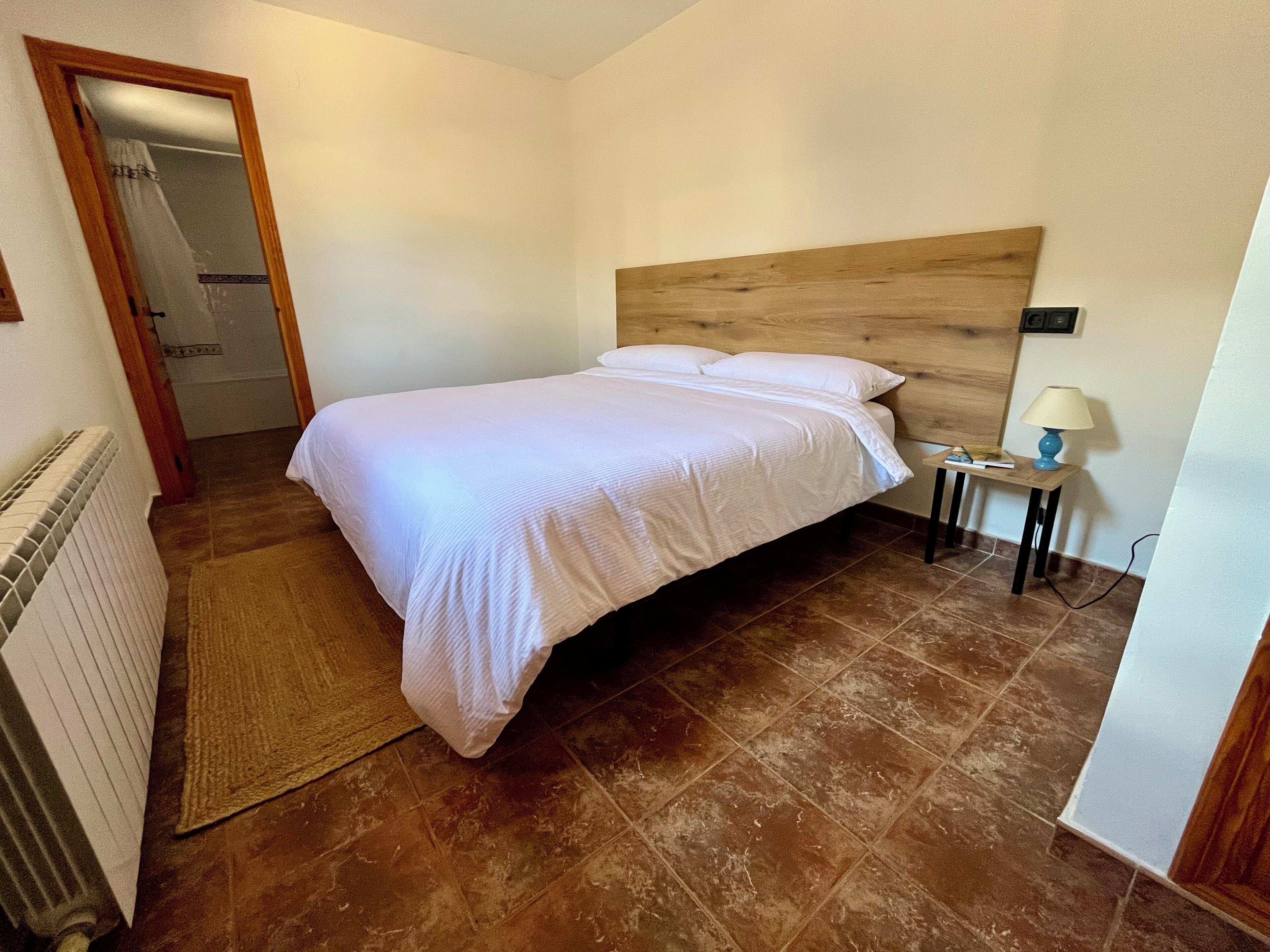 Quarto com cabeceira de madeira, cama com roupa de cama branca, mesa de cabeceira com abajur e radiador.