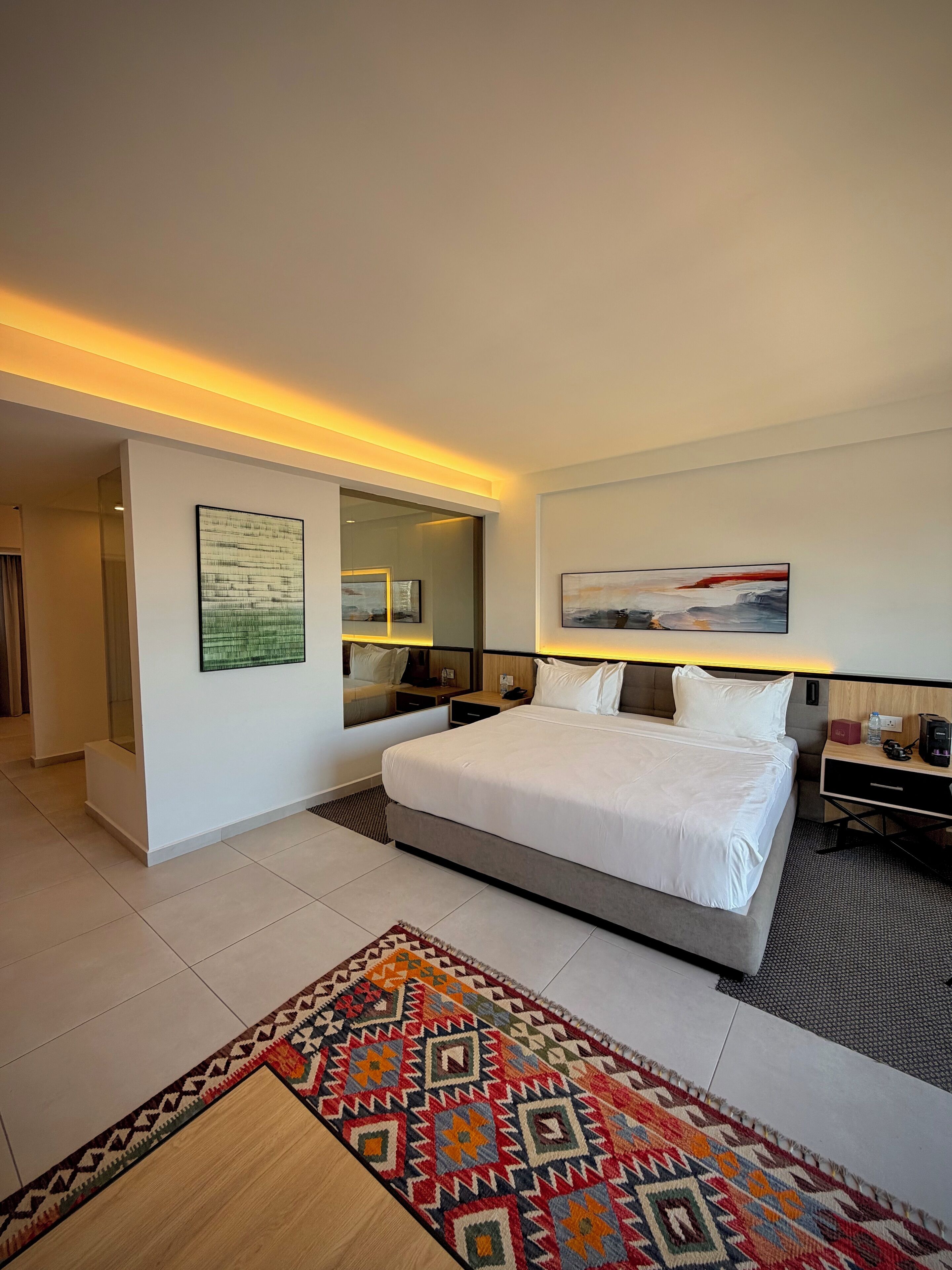 Quarto de hotel moderno com uma cama grande, uma pequena escrivaninha, uma mesinha de cabeceira e um espelho. Há um tapete colorido no chão e um quadro na parede.