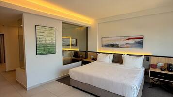 Quarto de hotel moderno com uma cama grande, uma pequena escrivaninha, uma mesinha de cabeceira e um espelho. Há um tapete colorido no chão e um quadro na parede.