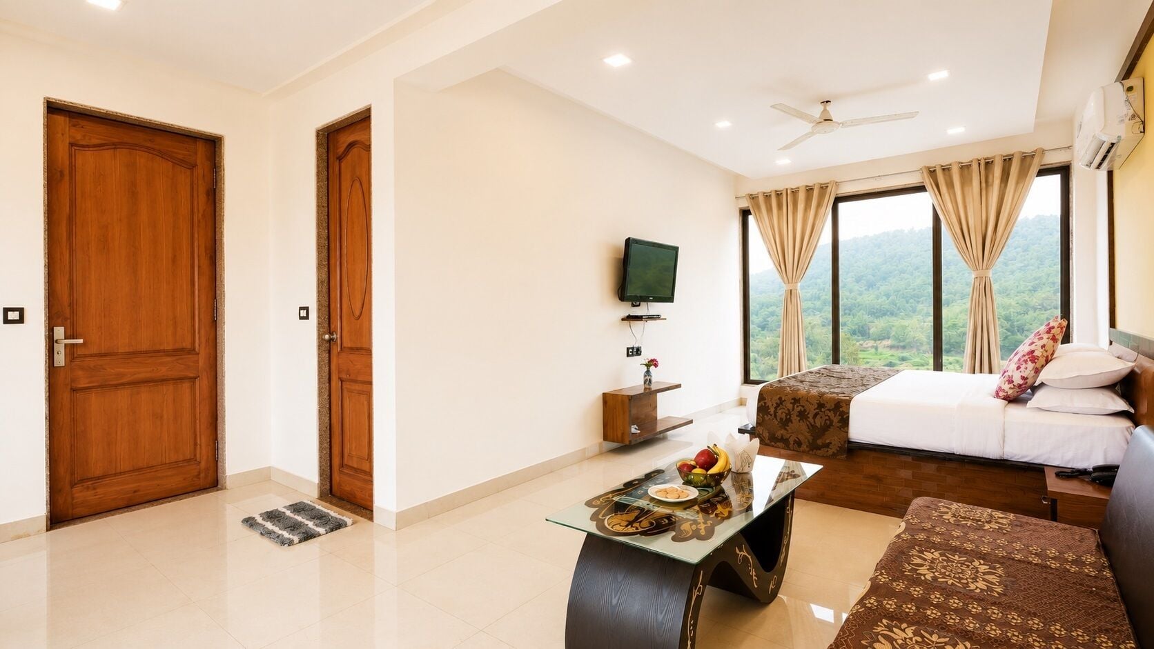 Quarto de hotel com cama, televisão, mesa de jantar com frutas, sofá e vista para o exterior.