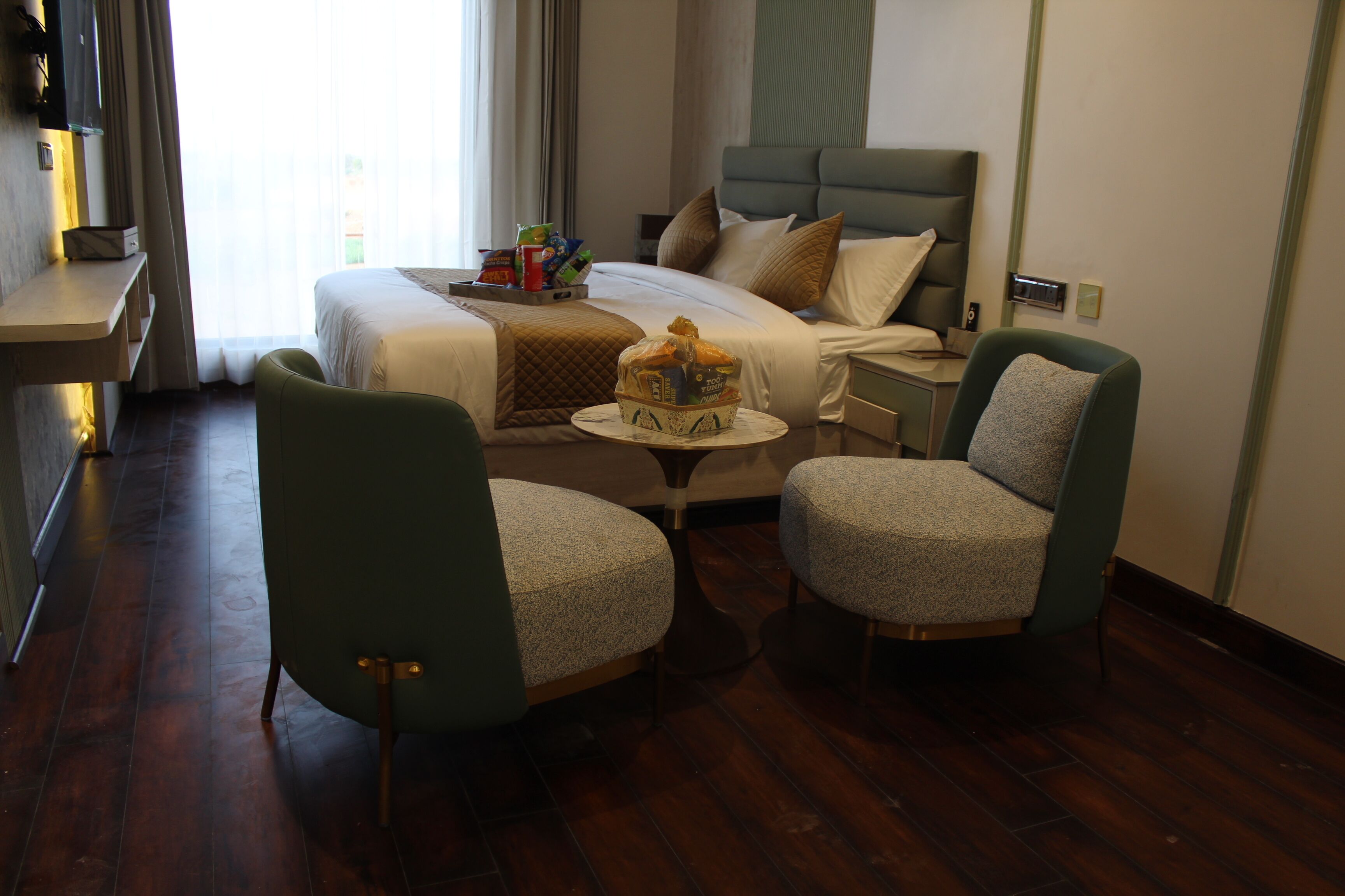 Quarto de hotel com uma cama grande, duas poltronas, uma mesinha redonda com petiscos e uma mesa de cabeceira com abajur.