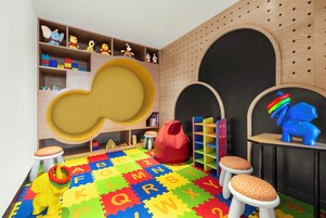 Um espaço de brincadeiras infantil com um chão de espuma colorido e com letras do alfabeto, estantes de madeira com brinquedos e uma ampla área de lazer circular.