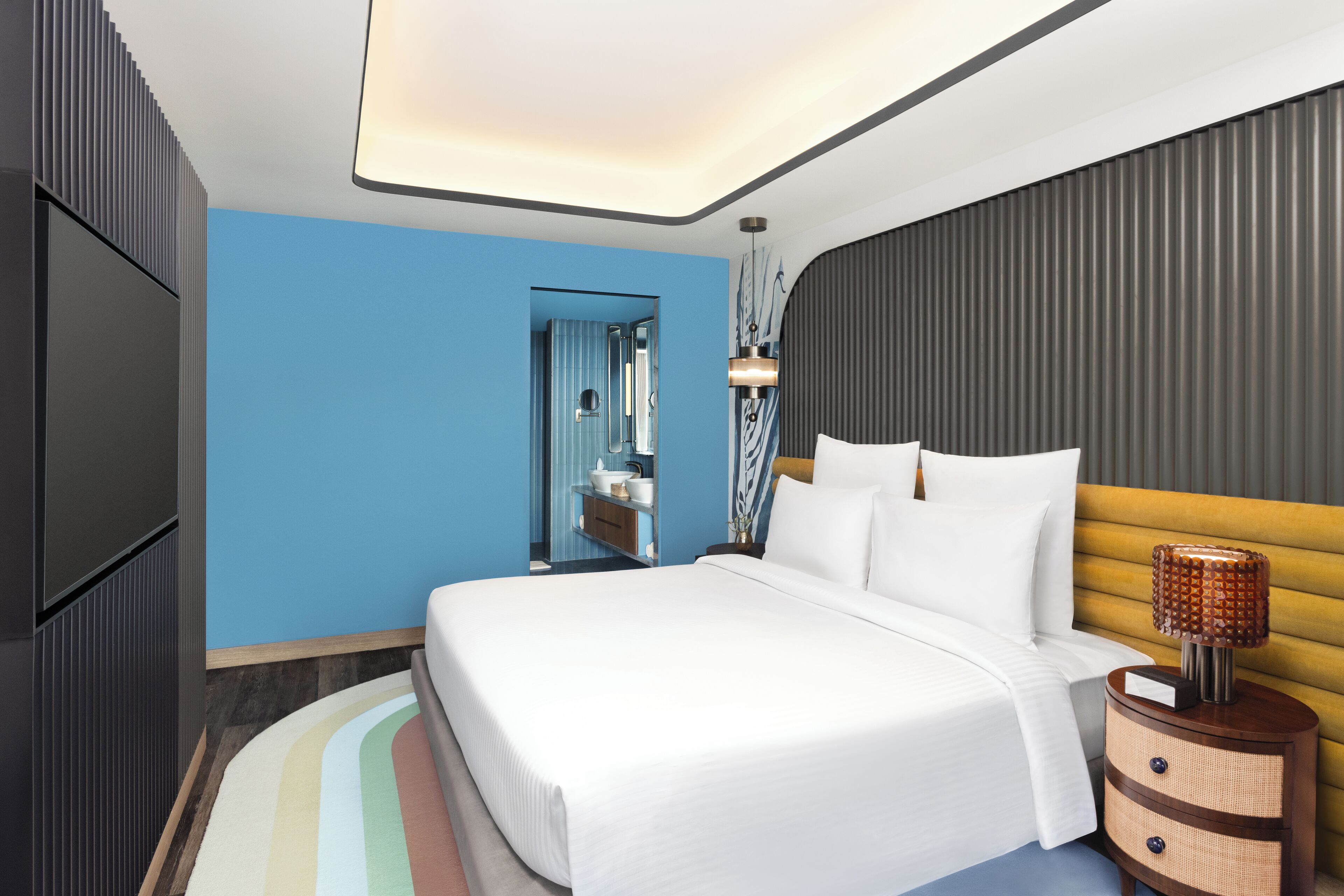 Quarto de hotel moderno com uma cama grande, uma televisão de tela plana, uma mesa de cabeceira com abajur e um banheiro visível através de uma porta aberta.