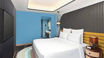 Quarto de hotel moderno com uma cama grande, uma televisão de tela plana, uma mesa de cabeceira com abajur e um banheiro visível através de uma porta aberta.