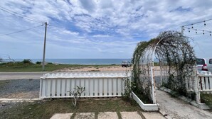 Vista costeira com cerca branca, uma pequena árvore e um carro estacionado perto da praia.