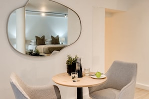 Quarto moderno com uma mesa redonda, duas poltronas cinzentas, um espelho, uma cama com almofadas estampadas, uma planta em vaso e uma maçã verde sobre um prato.
