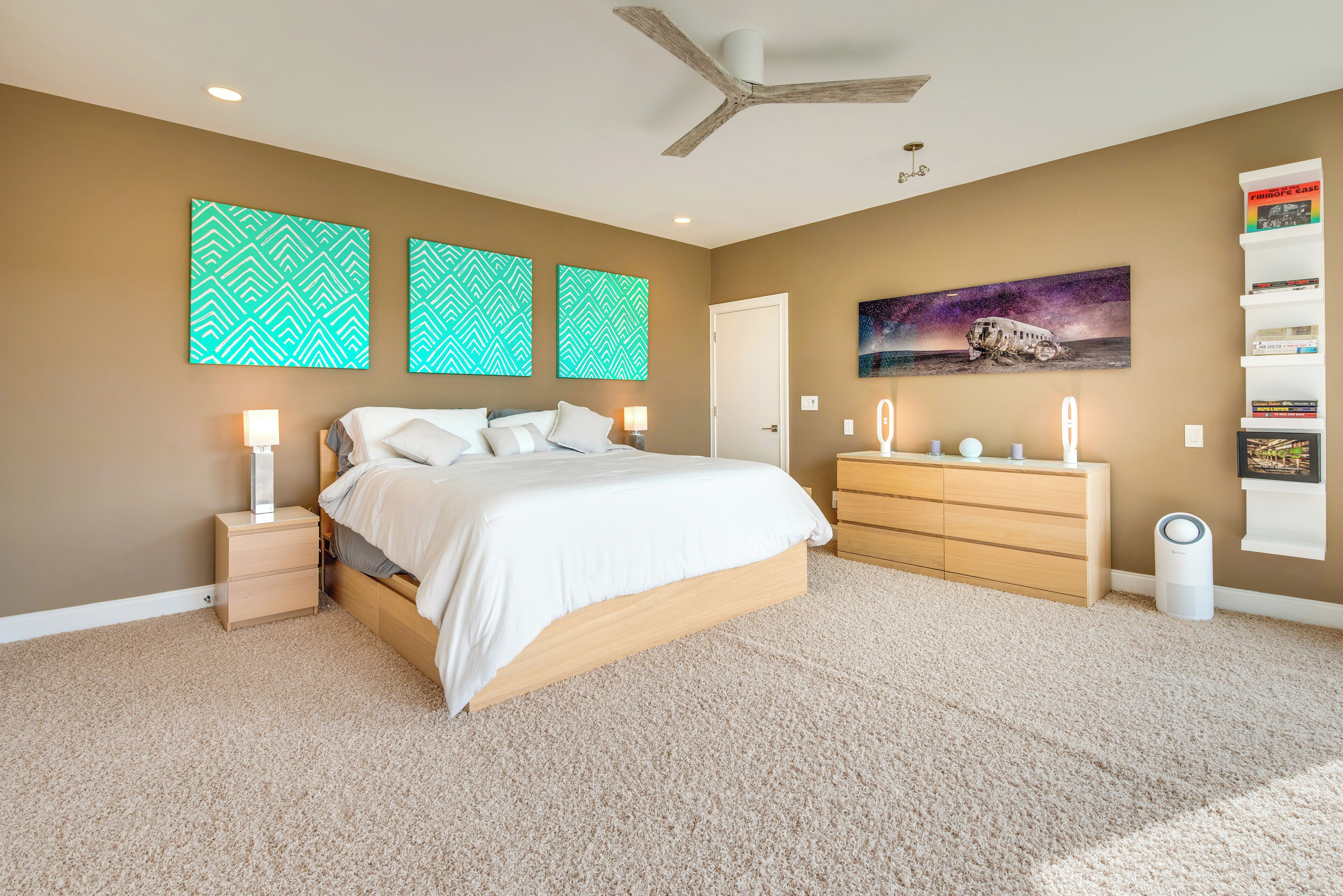Quarto com cama, mesas de cabeceira, cômoda, ventilador de teto e quadros nas paredes.