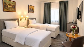 Quarto de hotel com duas camas, uma escrivaninha, uma cadeira, um vaso com flores e obras de arte nas paredes.