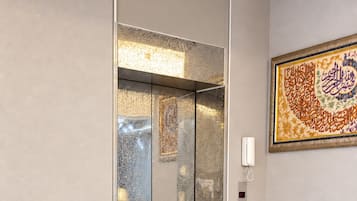 Um elevador com porta de vidro, telefone fixado na parede e um quadro com caligrafia.