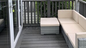 Uma área de estar coberta ao ar livre, com um banco e assento acolchoado, uma porta de vidro e um deck de madeira.