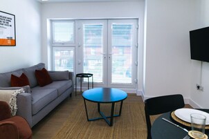 Uma sala de estar com um sofá cinza, uma mesa redonda azul, uma mesa pequena preta, uma cadeira preta e uma mesa de jantar com um copo e um prato.