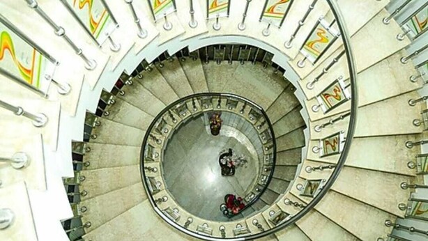 Uma escada em espiral com um vão central aberto, cercada por corrimãos com desenhos coloridos.
