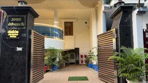 Uma entrada com portão, placa, plantas em vasos e uma galeria coberta que leva a um edifício.
