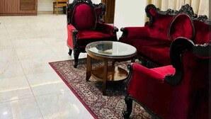 Quarto com cadeiras estofadas em vermelho, uma mesa redonda pequena, tapete estampado, armário de madeira e porta com cortinas.
