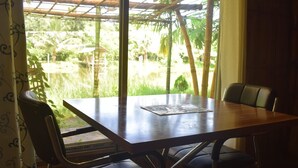 Uma mesa de madeira com um jogo americano, duas cadeiras e vista para o verde através de uma janela.