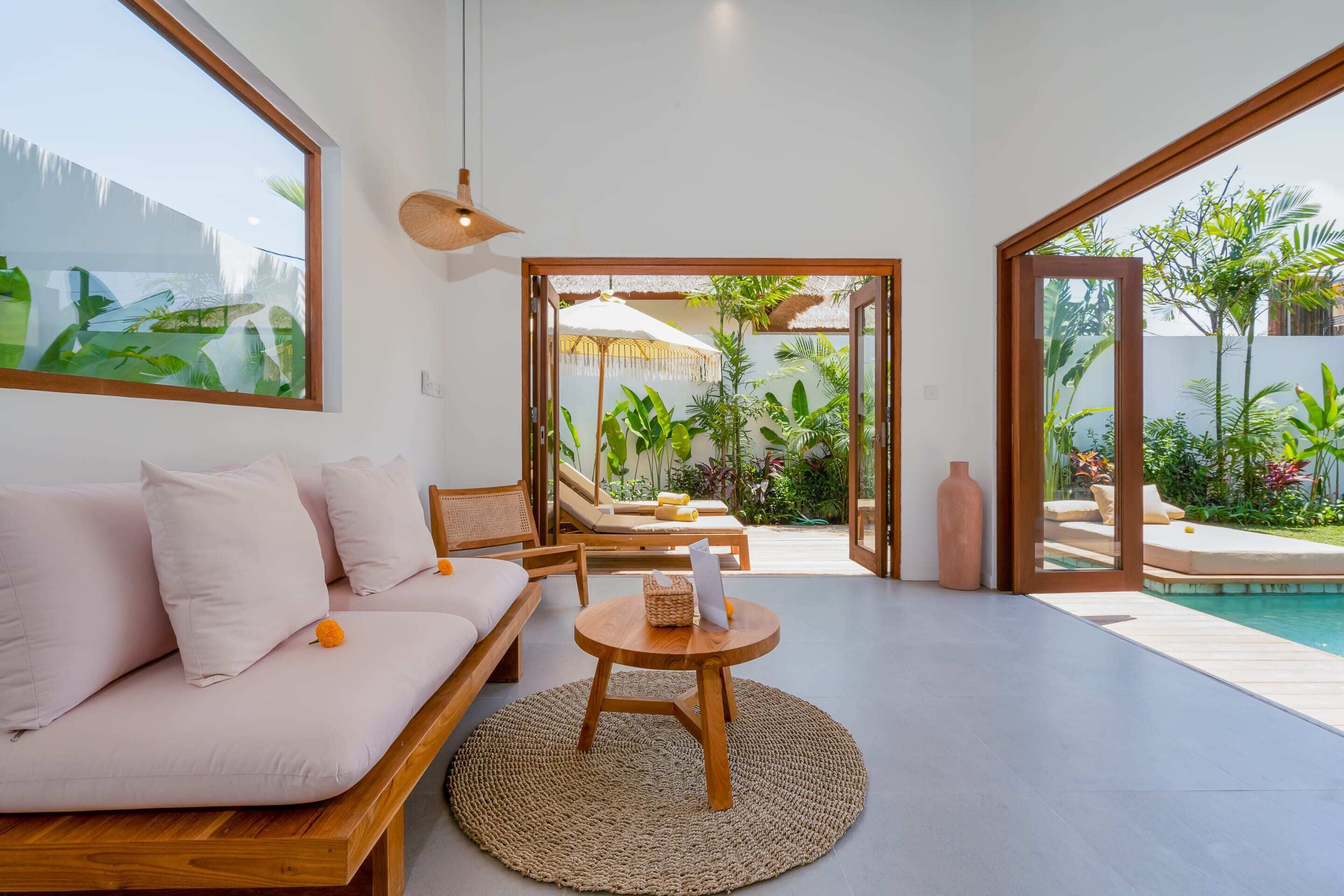 Uma sala de estar moderna com sofá, cadeiras, uma mesa redonda de madeira e vista para uma área de piscina externa.