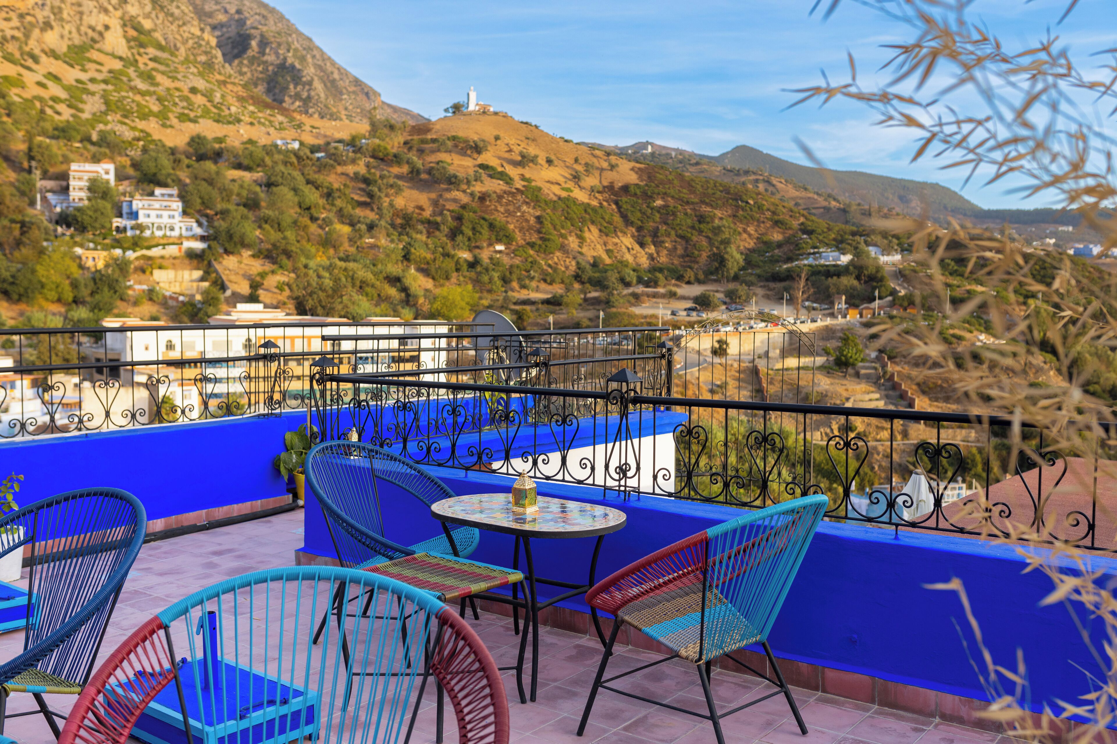 Um terraço com cadeiras de vime azuis e vermelhas, uma mesa redonda e vista para uma colina com edifícios e um farol.