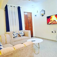 Uma sala de estar com um sofá bege, uma mesa de centro redonda, uma televisão na parede e uma porta ao fundo.