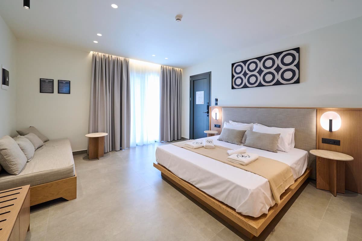 Quarto de hotel moderno com uma cama grande, mesas de cabeceira, um sofá e vista para o exterior.