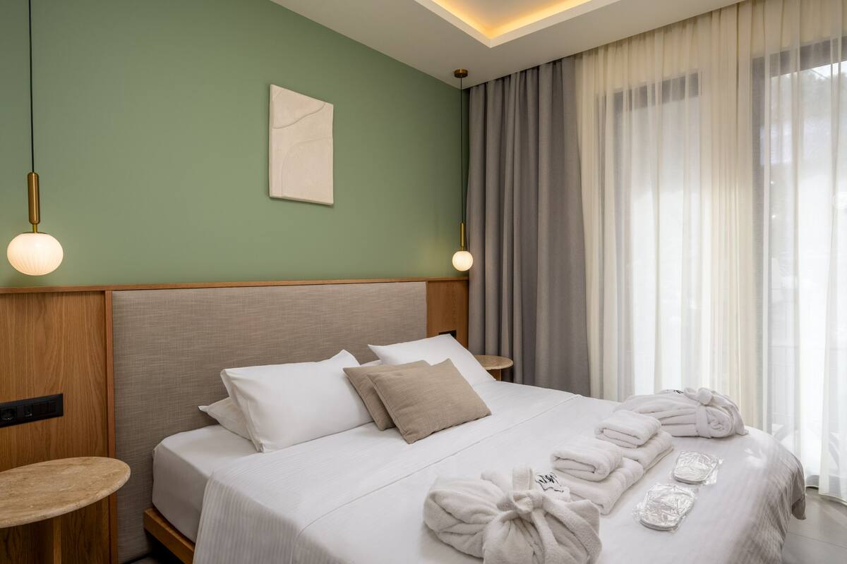 Cama bem arrumada com lençóis brancos e travesseiro bege, mesa de cabeceira com abajur e janela com cortinas finas.