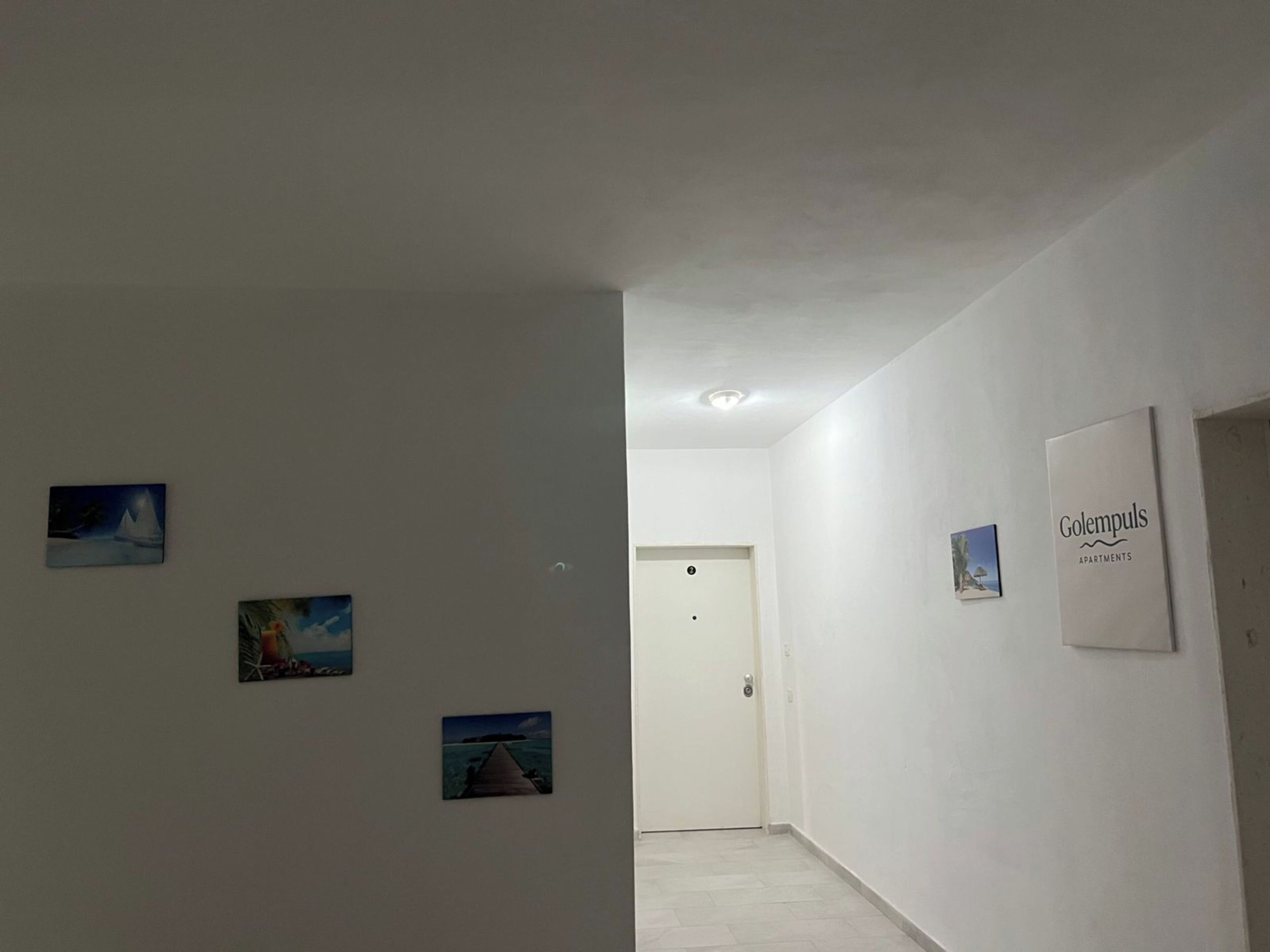 Um corredor com paredes brancas, uma porta no final e três quadros na parede esquerda.
