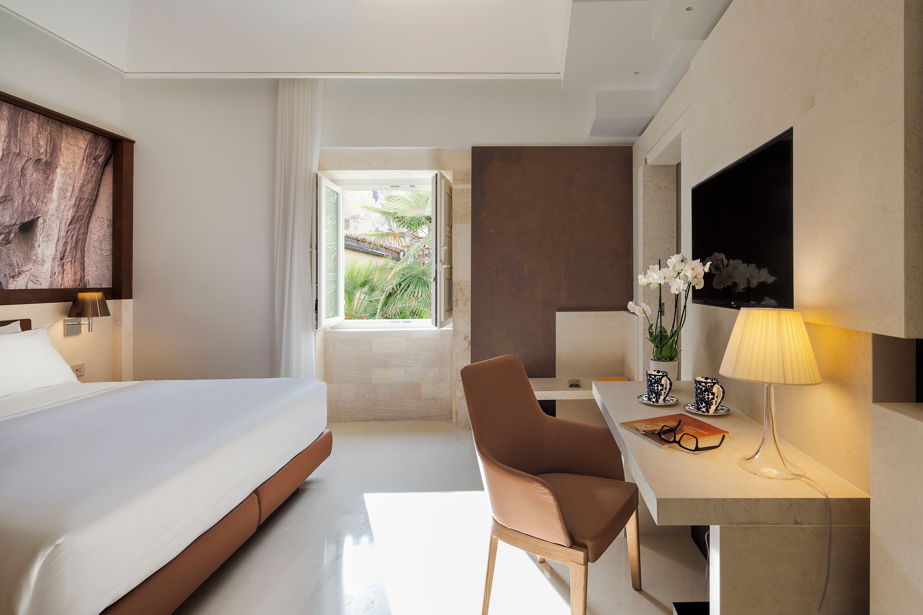 Quarto de hotel moderno com cama, escrivaninha, cadeira, luminária e uma grande janela com vista para áreas verdes.