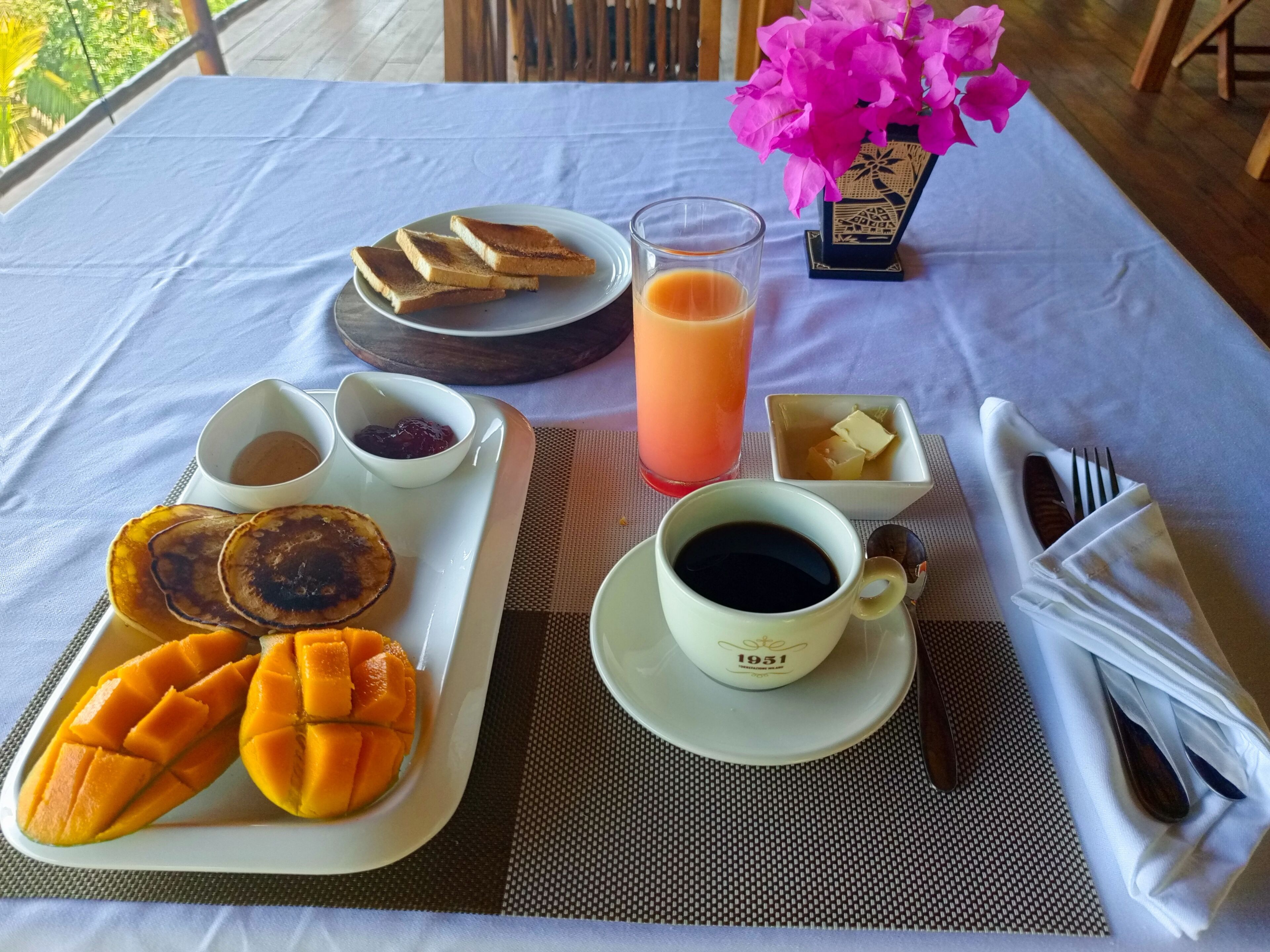 Uma mesa de café da manhã com torradas, panquecas, uma xícara de café, um copo de suco de laranja e uma tigela de frutas.