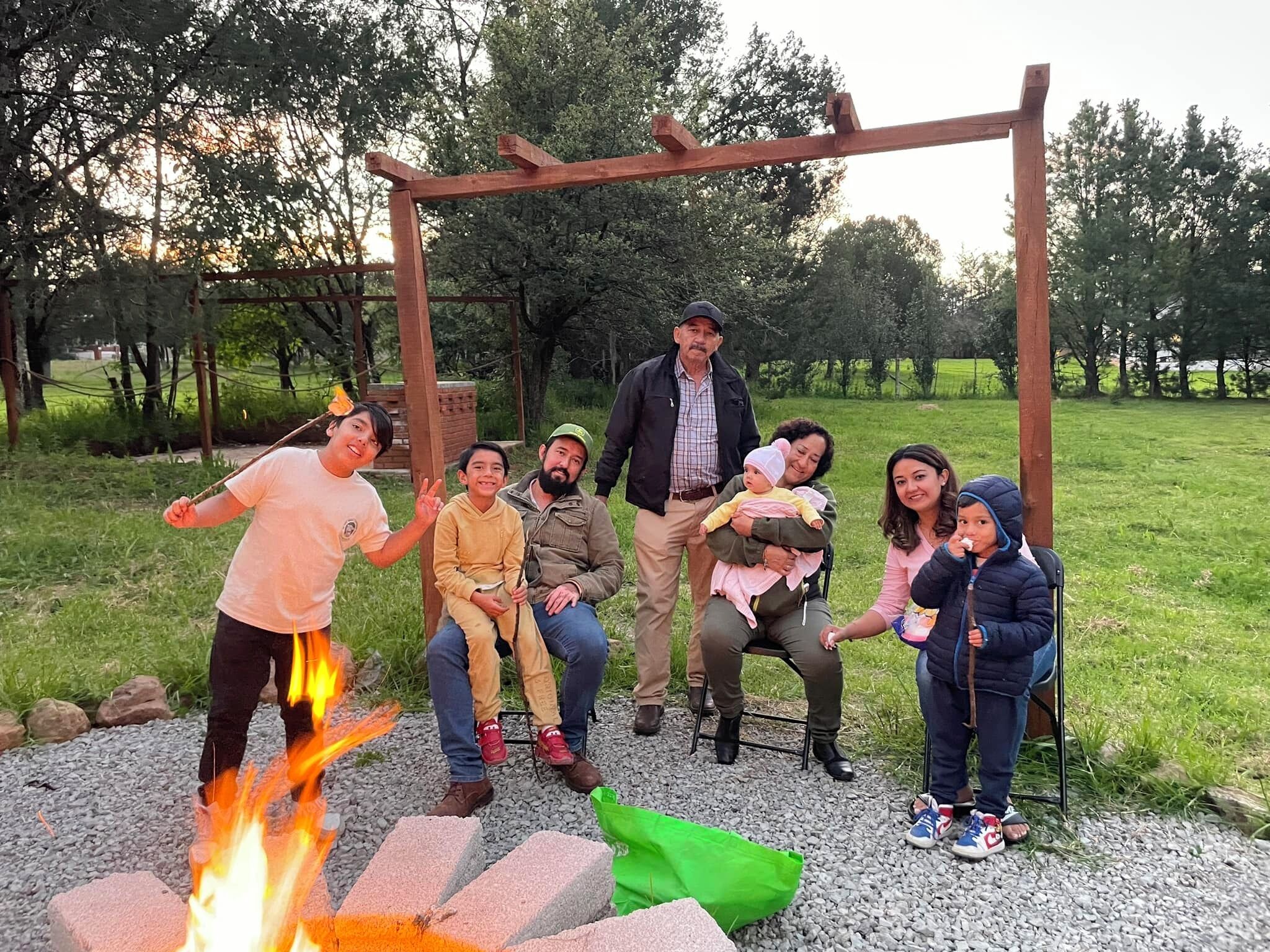 Um grupo de pessoas reunidas ao redor de uma fogueira em ambiente externo, com uma estrutura de madeira acima.