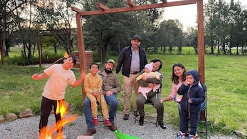 Um grupo de pessoas reunidas ao redor de uma fogueira em ambiente externo, com uma estrutura de madeira acima.