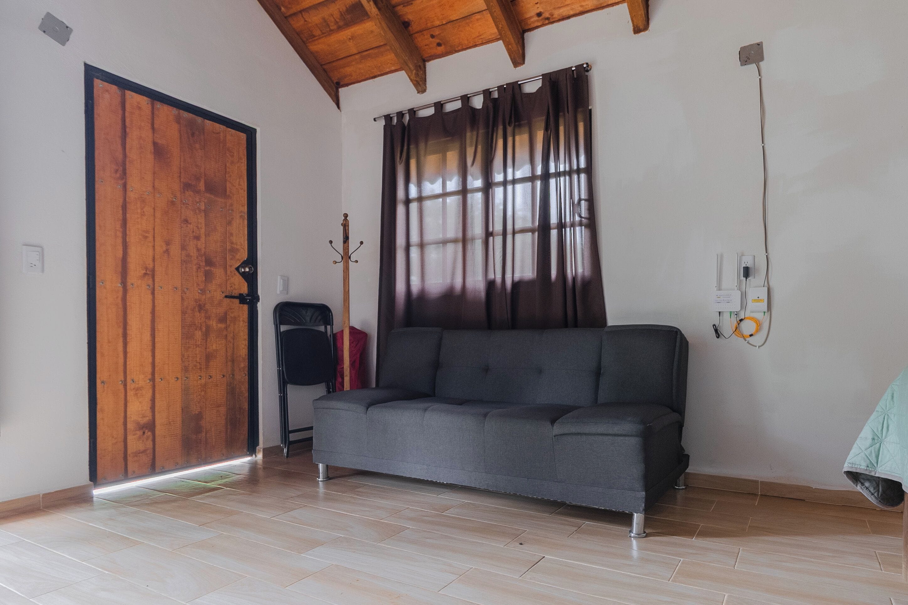 Quarto com porta de madeira, sofá cinza, guitarra e tomada na parede.