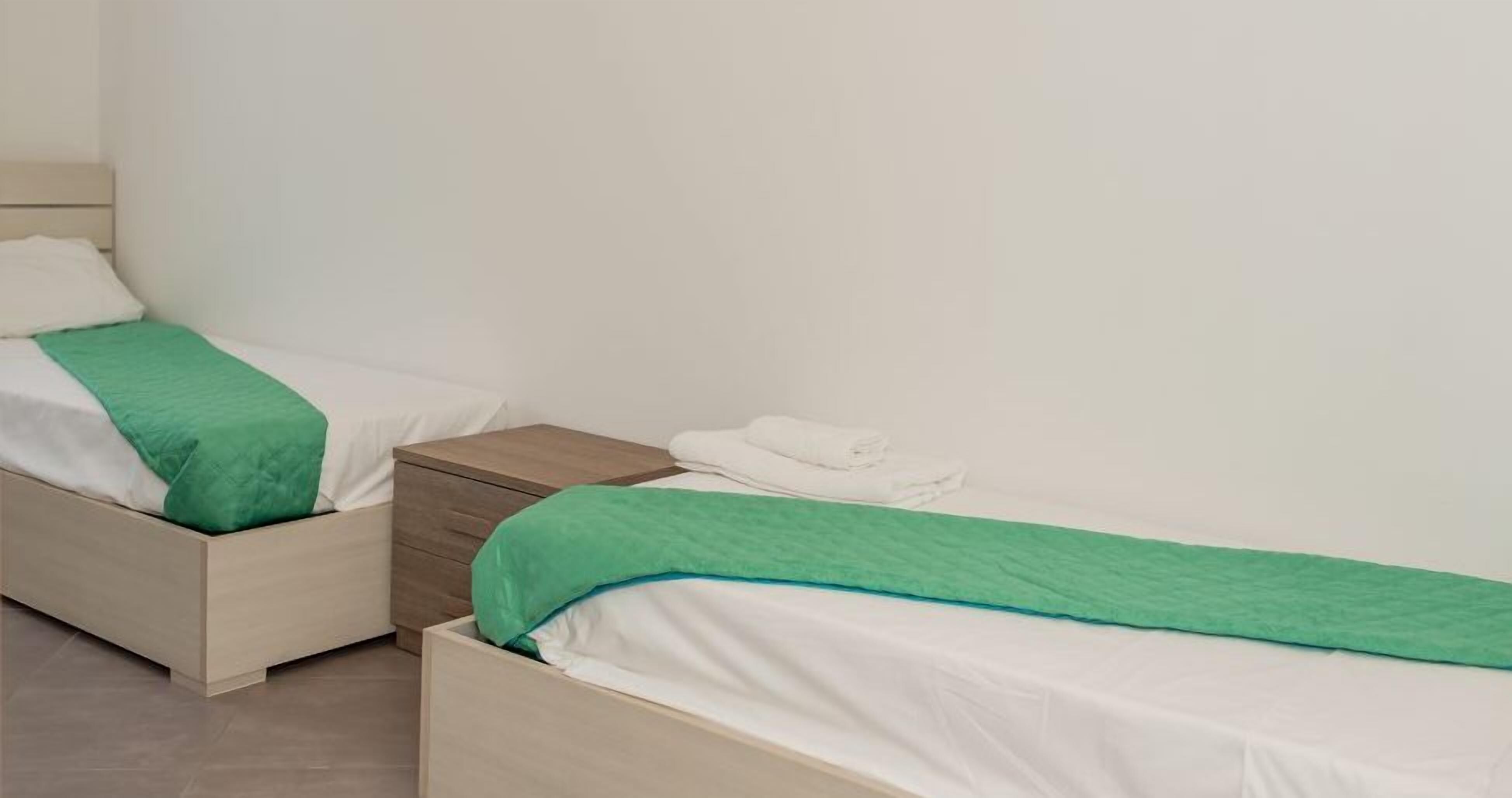 Duas camas de solteiro com roupa de cama verde, um criado-mudo de madeira e uma toalha branca sobre ele.
