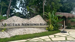 Placa indicando Mactan Newtown Beach, com muro de pedra e vegetação tropical.