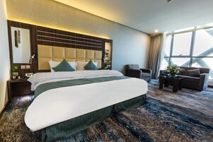 Quarto de hotel moderno com uma cama grande, cabeceira com painel iluminado, área de estar com sofá e poltronas, e uma janela ampla com vista.