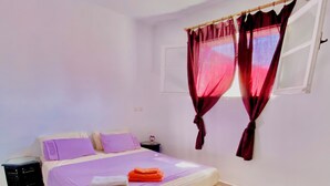 Um quarto com cama, almofadas, mesa de cabeceira e janela com cortinas.