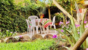 Um jardim com mesa e cadeiras, cercado por vegetação e flores.