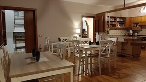 Uma sala de jantar com mesa de madeira e cadeiras brancas, uma cozinha com armários de madeira e bancada, e uma escada que leva a um andar superior.