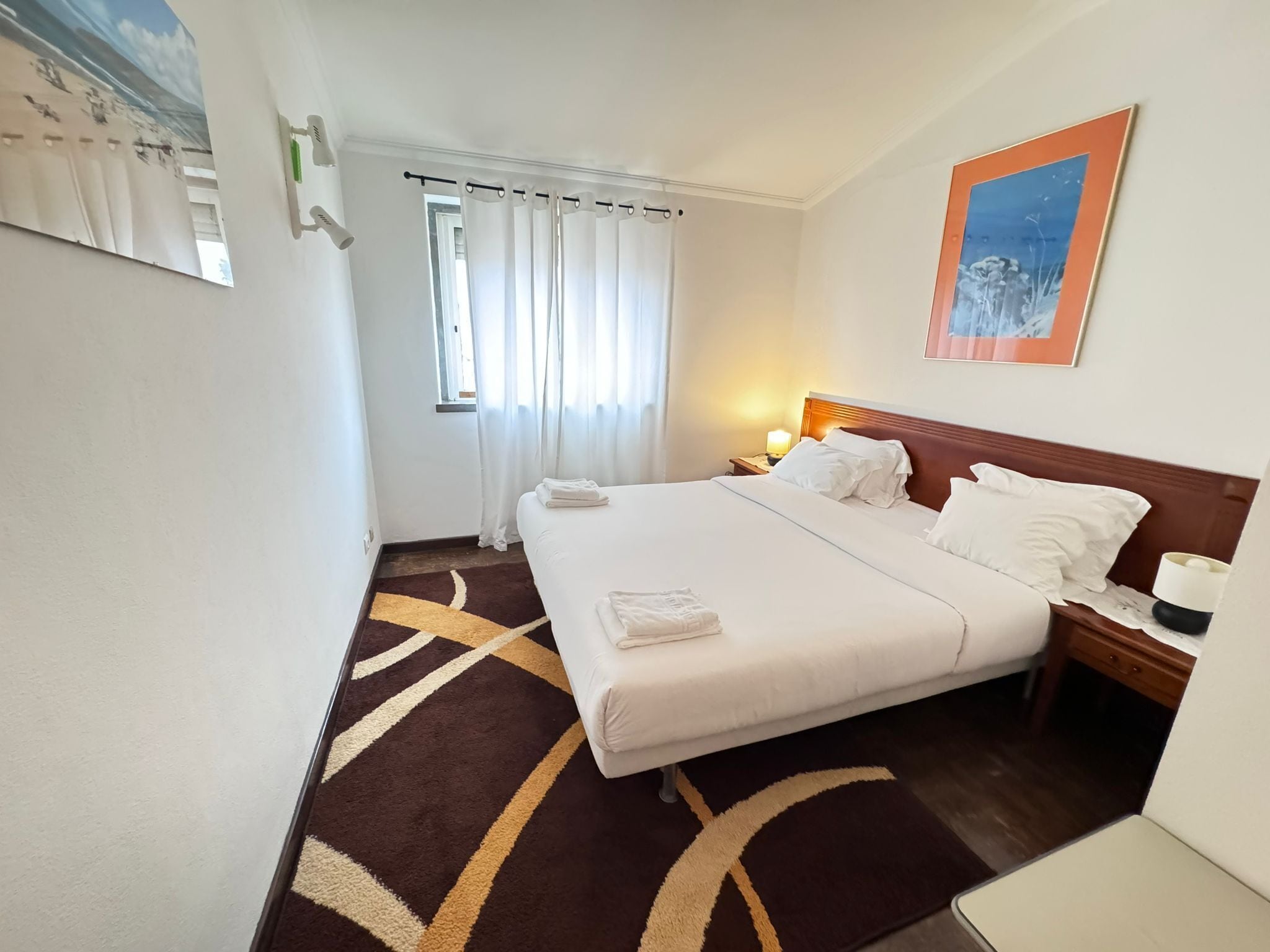 Quarto de hotel com uma cama grande, mesa de cabeceira com abajur, janela com cortinas e um quadro na parede.