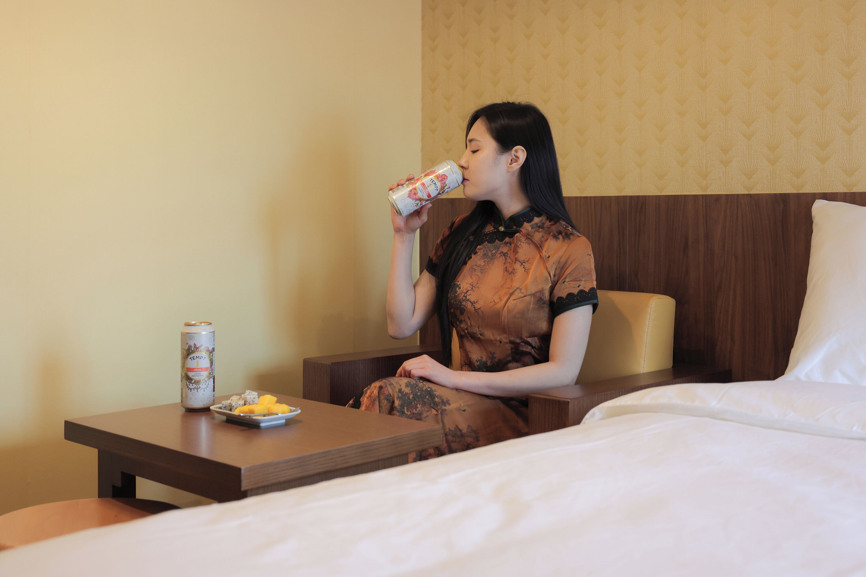 Uma mulher sentada em uma cama em quarto de hotel, segurando uma lata. Há um prato com comida e uma garrafa na mesinha de cabeceira.