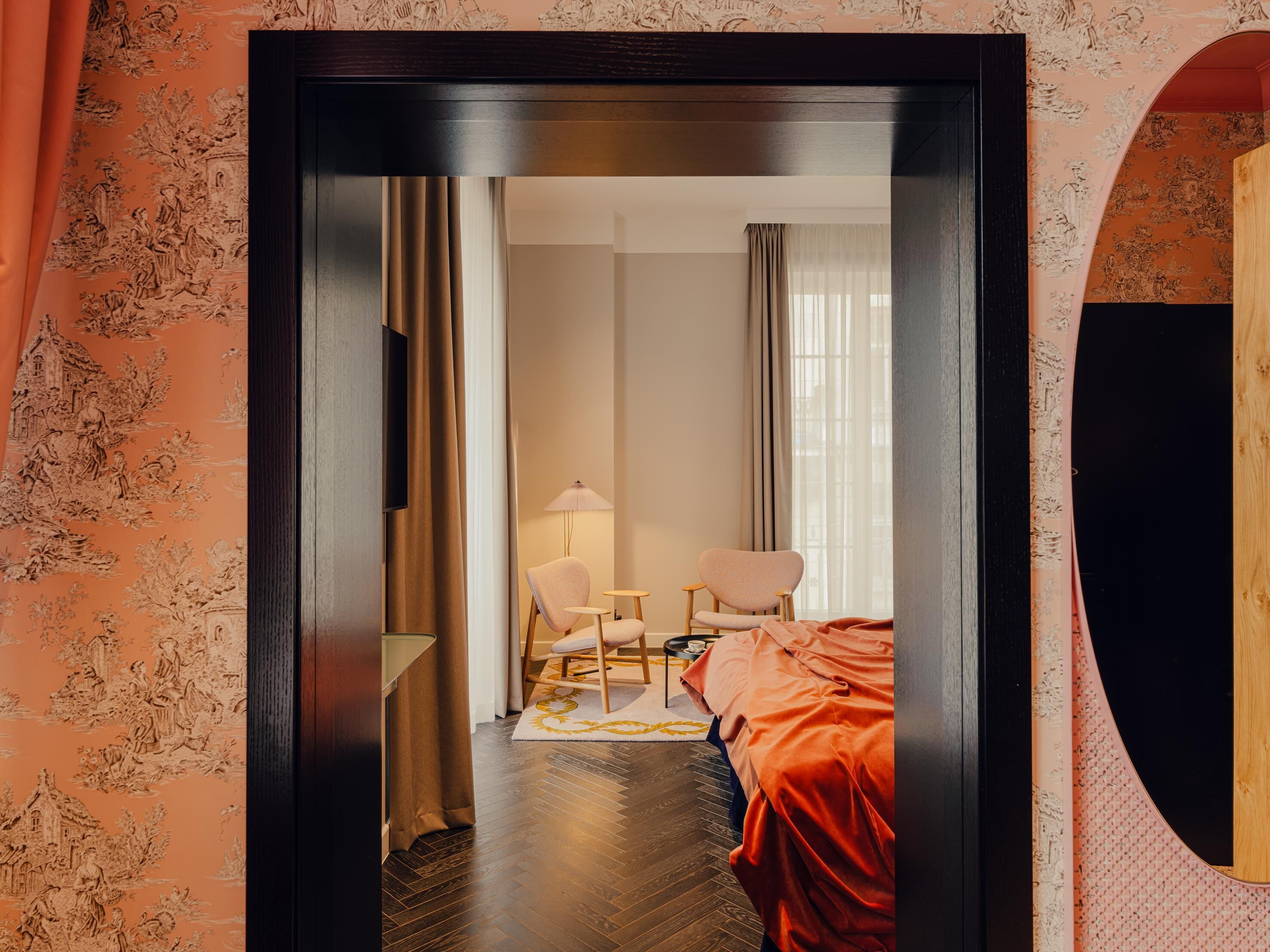 Quarto de hotel com piso de madeira, cama com cobertura vermelha, duas cadeiras, um abajur e uma TV na parede.