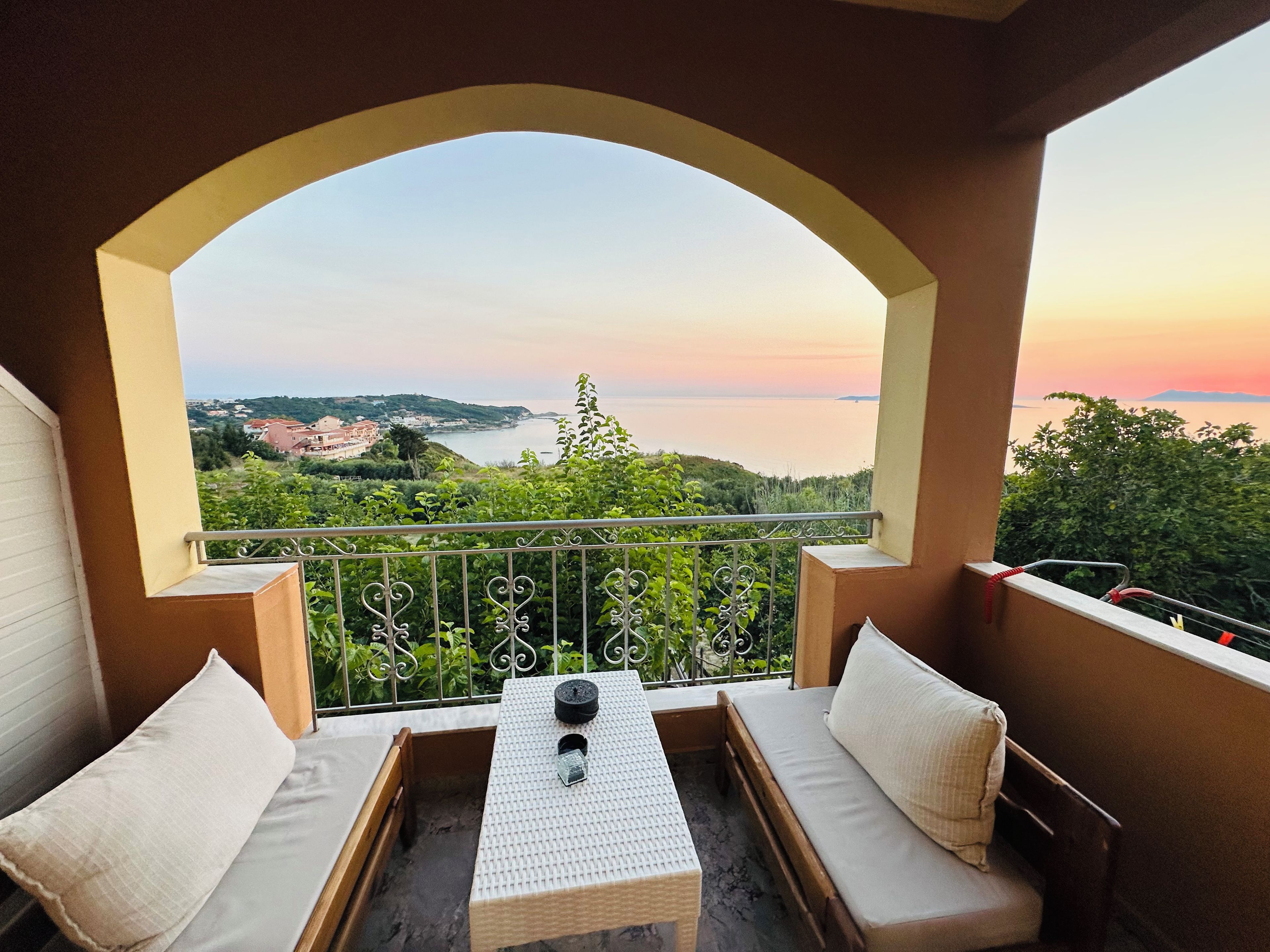 Varanda com vista para o mar e colinas, mobiliada com uma mesa e dois sofás.