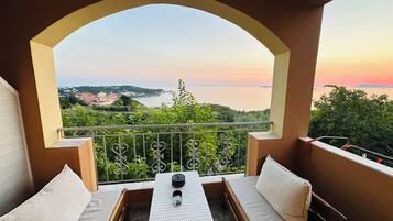 Varanda com vista para o mar e colinas, mobiliada com uma mesa e dois sofás.