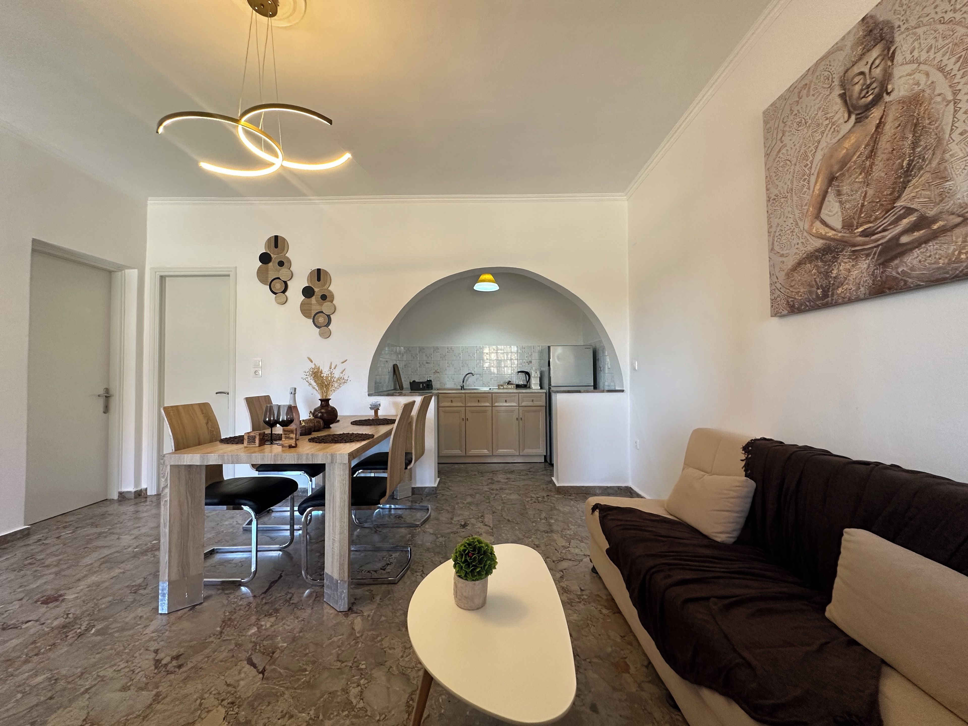 Uma sala moderna com sofá, mesa de centro e área de jantar com cadeiras e mesa. Uma cozinha é visível através de um arco.