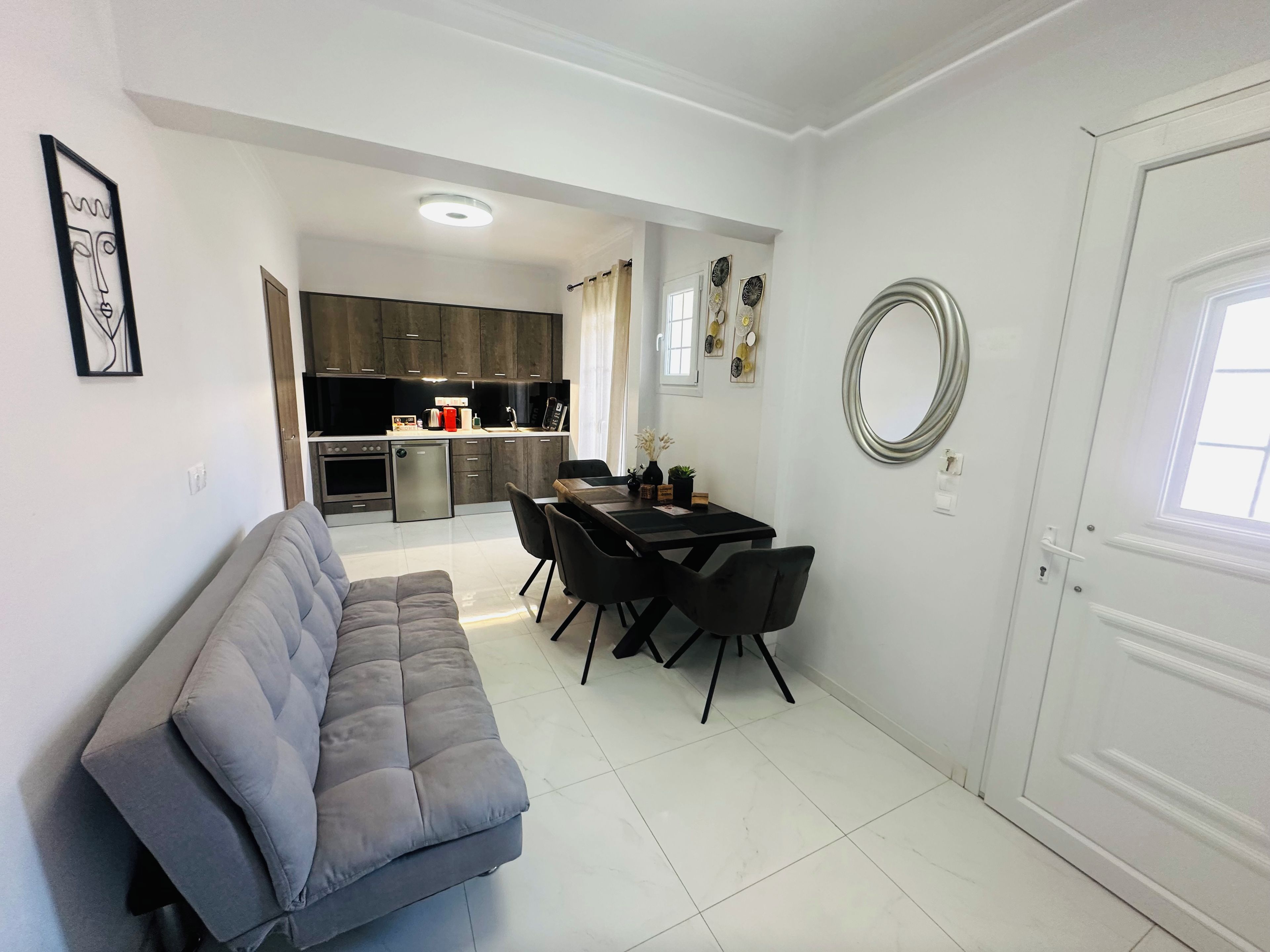 Um apartamento moderno com um sofá cinzento, uma mesa de jantar com cadeiras, uma cozinha com armários escuros e uma porta branca.